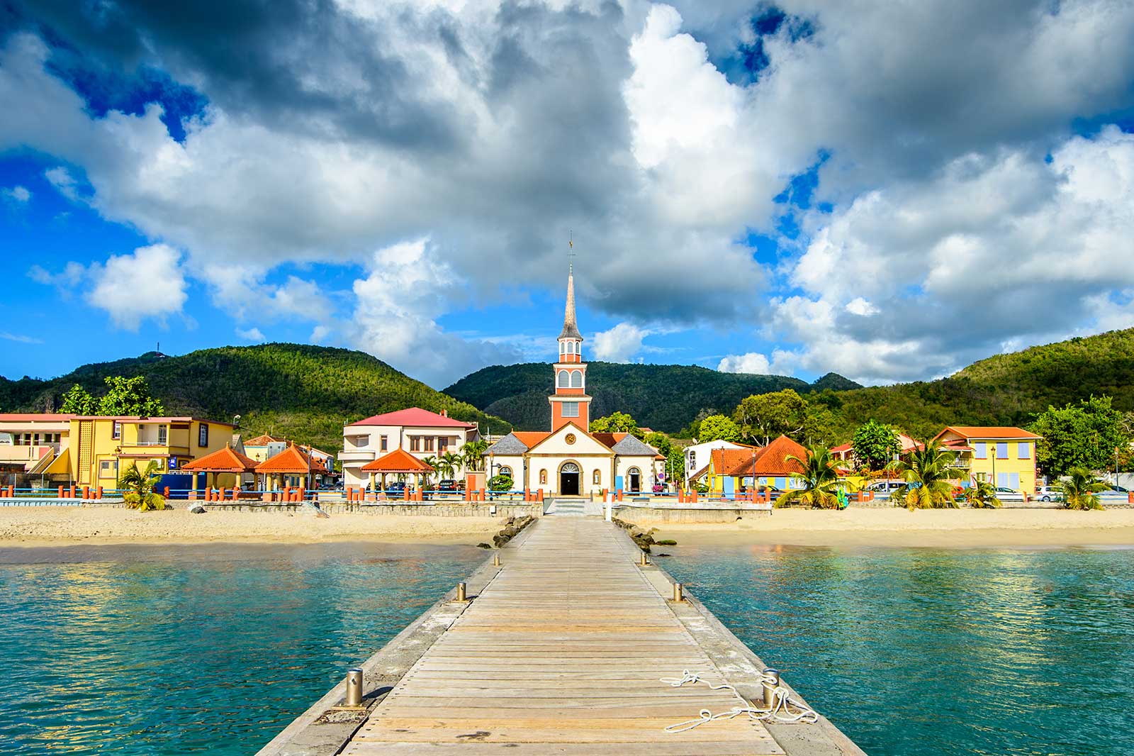 A view from a long pier to a small town with a church and colourful buildings by the beach.
