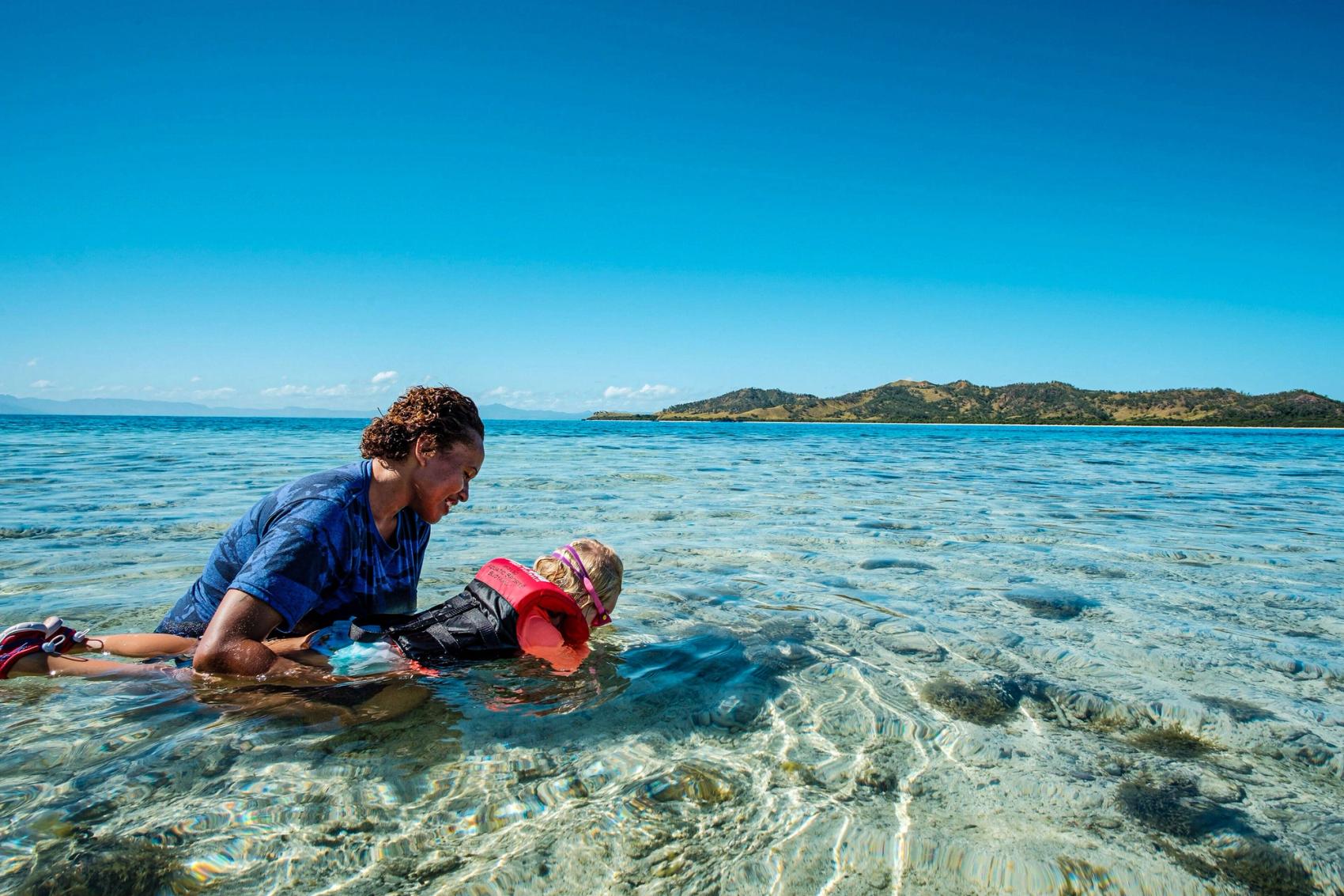 A woman holds a child wearing a life vest and snorkel mask over shallow clear water with an island in the distance.