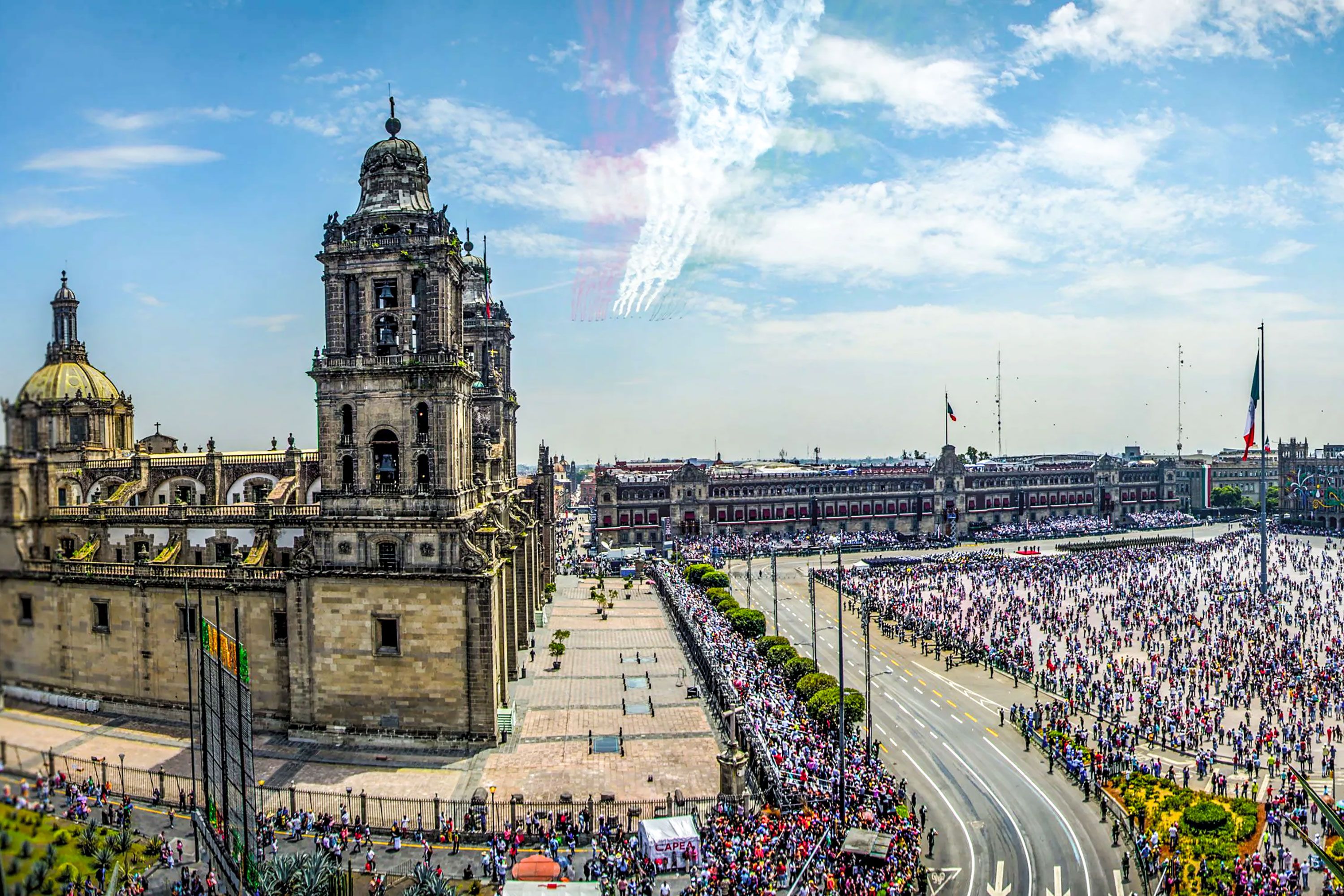 Panoramic view looking down into large crowded square with church and jets flying overhead.