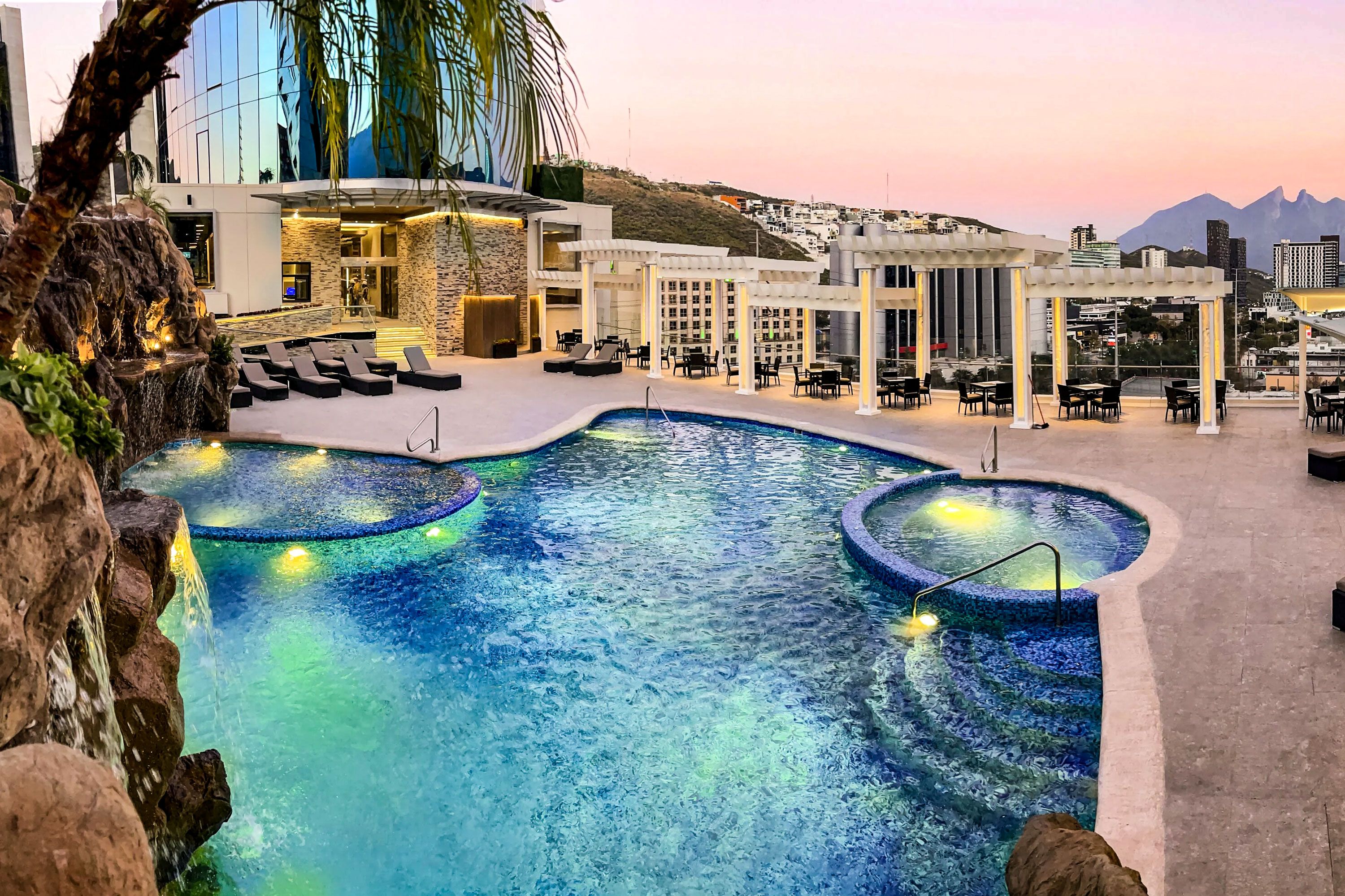 A swimming pool with built-in Jacuzzis and rock waterfalls surrounded by lounge chairs, pergolas, and a building entrance with city and mountain in the distance.