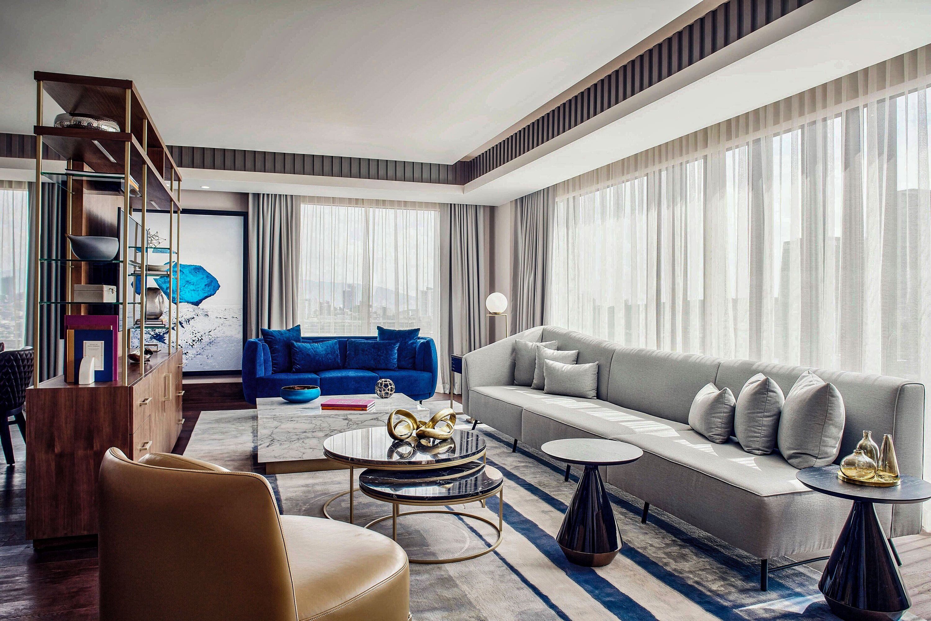 A corner room with modern couches and chairs and tables with carpet and translucent curtains.