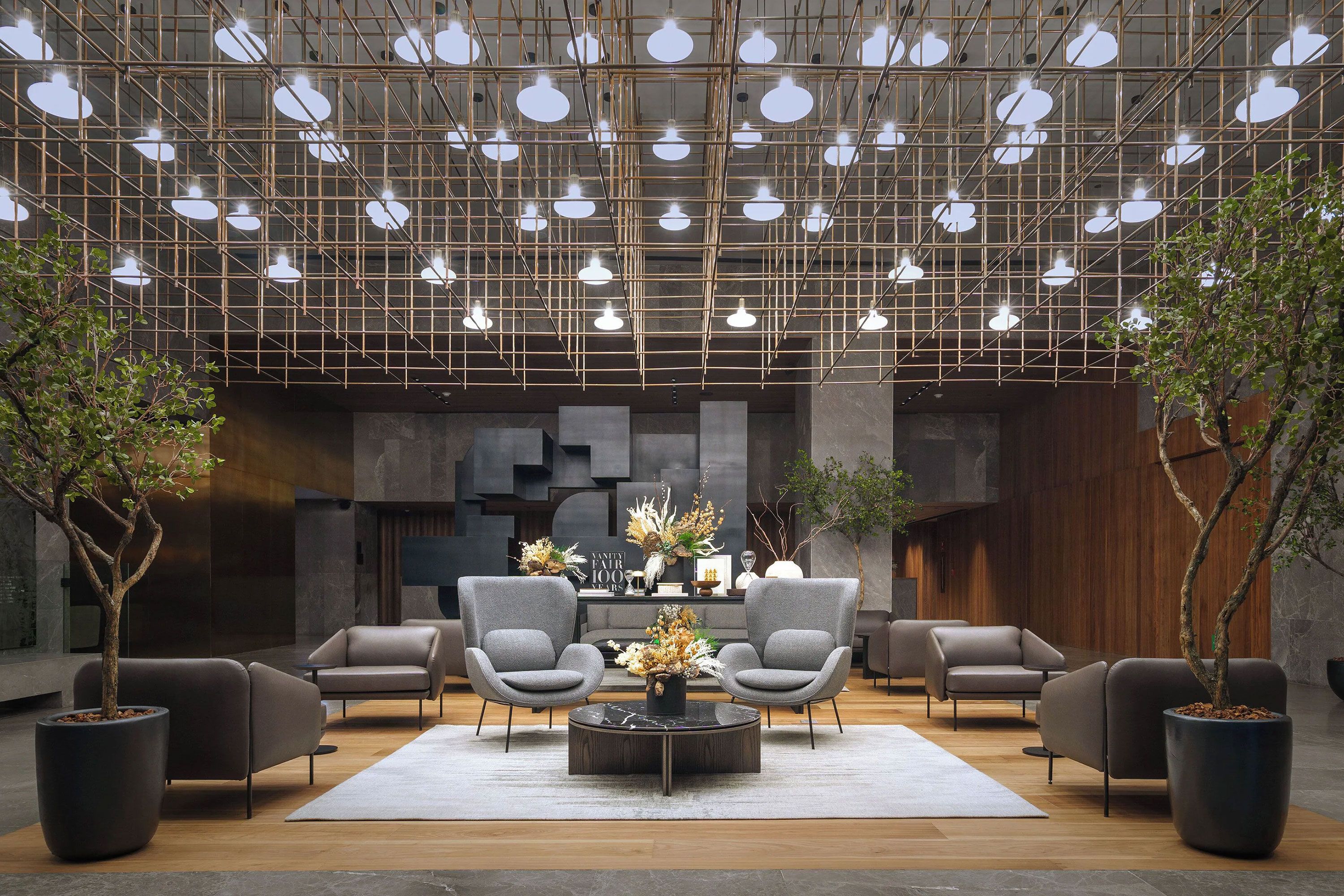 A lounge area with modern furnishings, potted trees, elaborate metal ceiling feature and grey stone floor and walls.